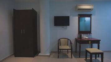 Quarto com guarda-roupa escuro, uma cadeira, uma mesa pequena com espelho e televisão na parede.