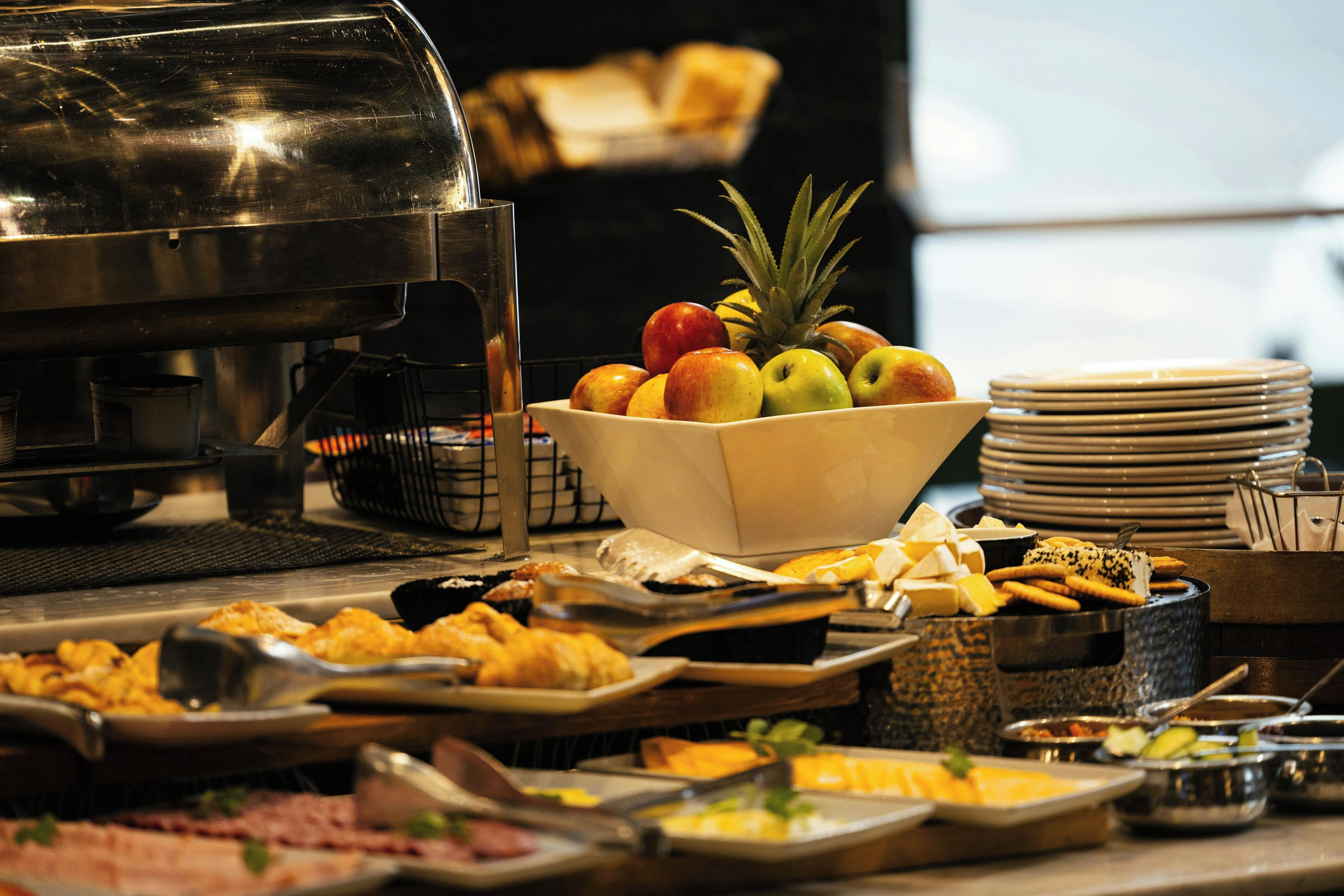 Disposição de buffet com diversos alimentos, incluindo frutas e queijos, e uma máquina de café ao fundo.
