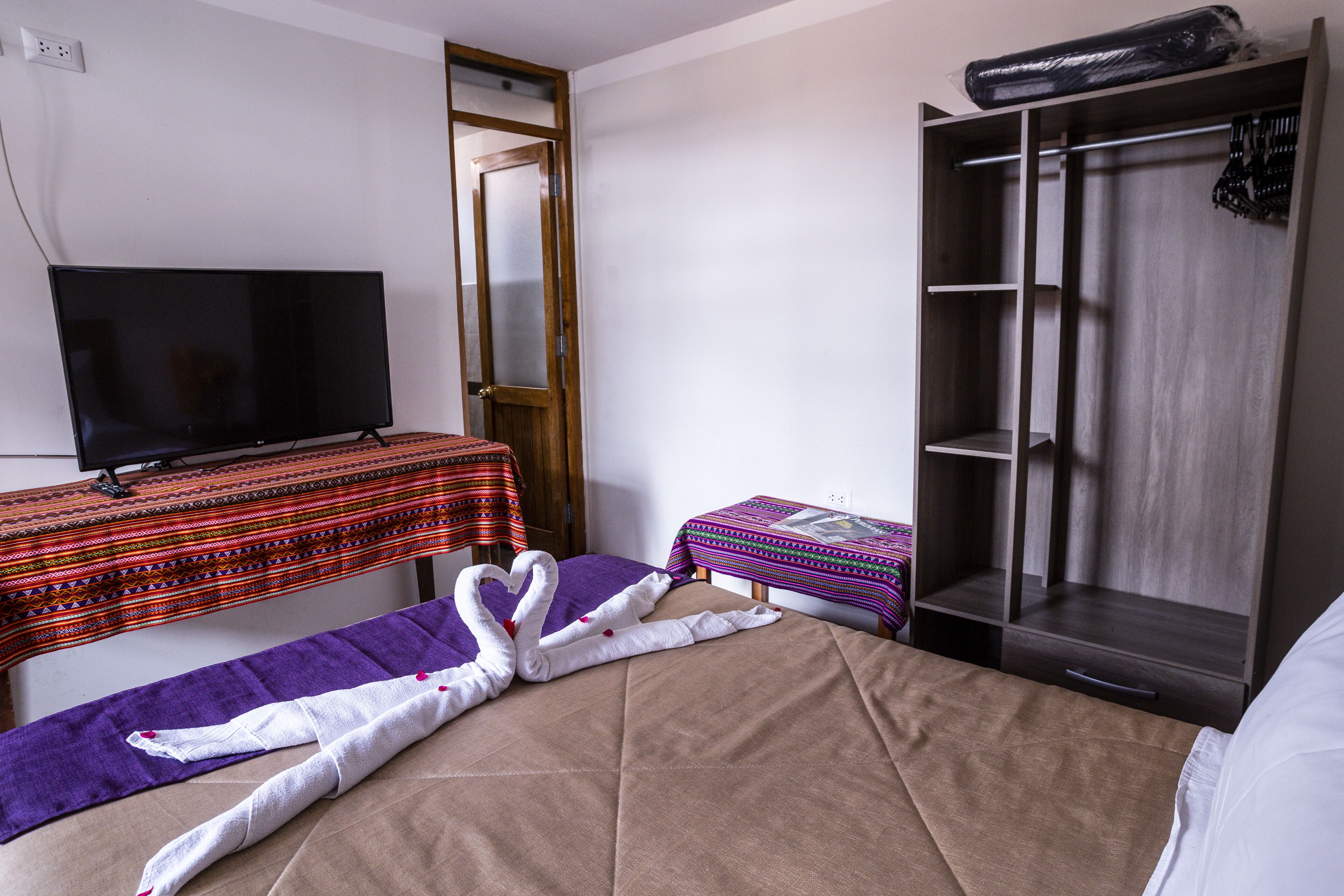 Quarto de hotel com cama, televisão, guarda-roupa e uma pequena mesa com toalha de mesa estampada.