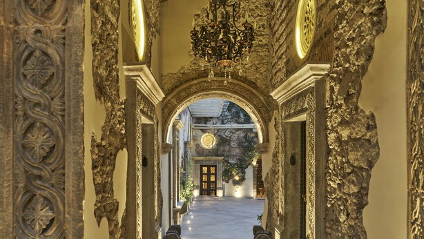 Um corredor ornamentado com esculturas elaboradas, um candelabro e uma porta no final.