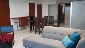 Uma sala de estar com sofá, mesa de jantar e cadeiras, e uma pequena cozinha.