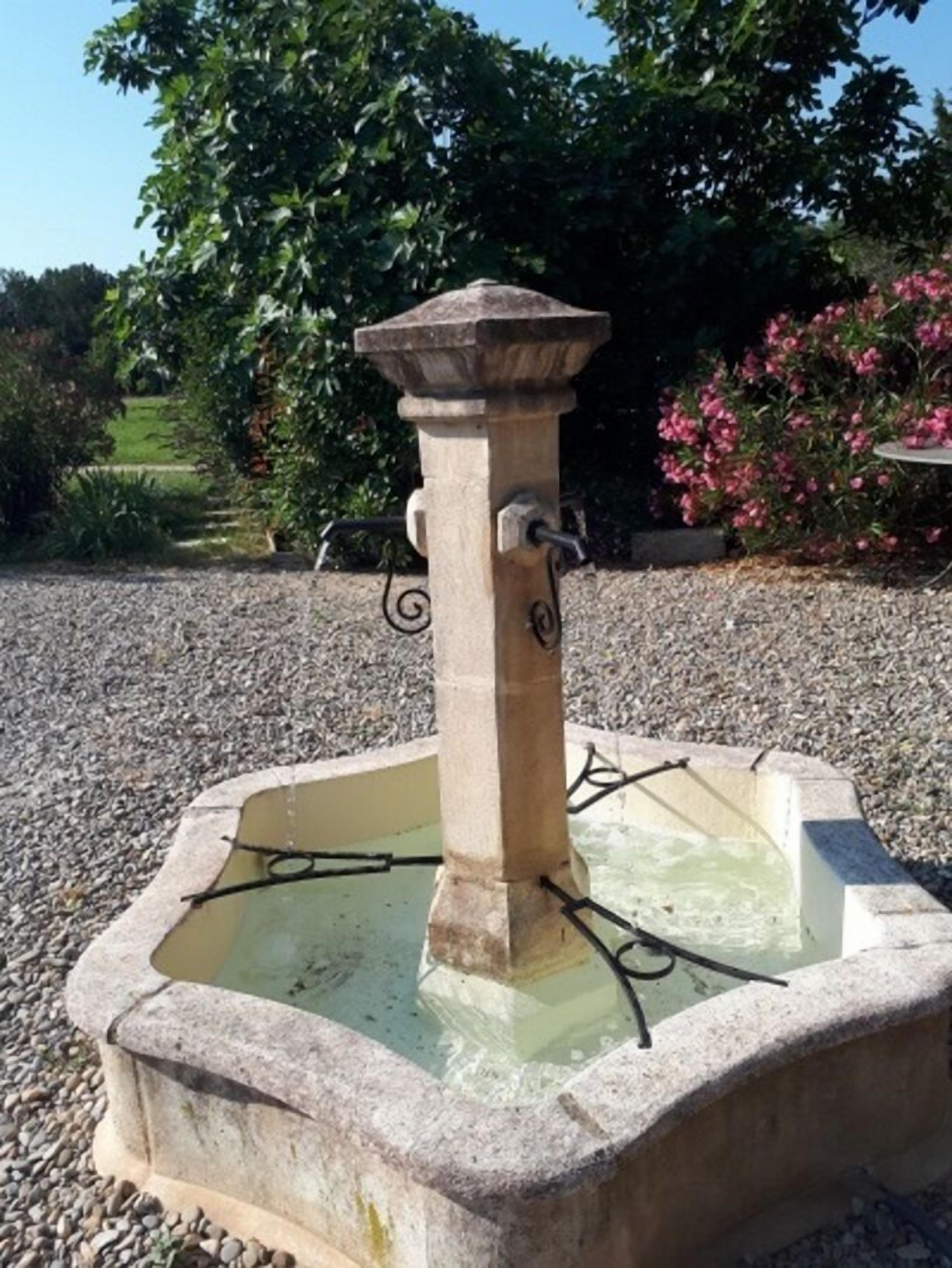 Uma fonte de pedra com bacia circular, rodeada por cascalho e vegetação.