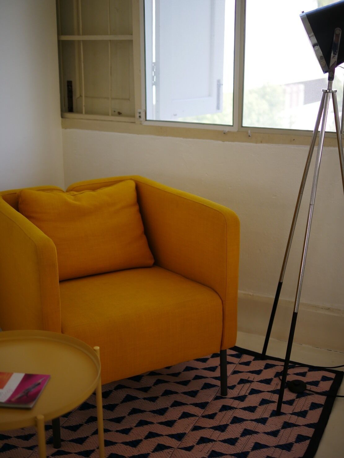 Quarto com sofá amarelo, uma mesa redonda pequena com uma revista, tapete estampado e abajur.