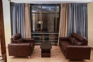 Uma sala de estar com dois sofás de couro marrom, uma mesinha de centro de madeira e uma janela grande com cortinas.
