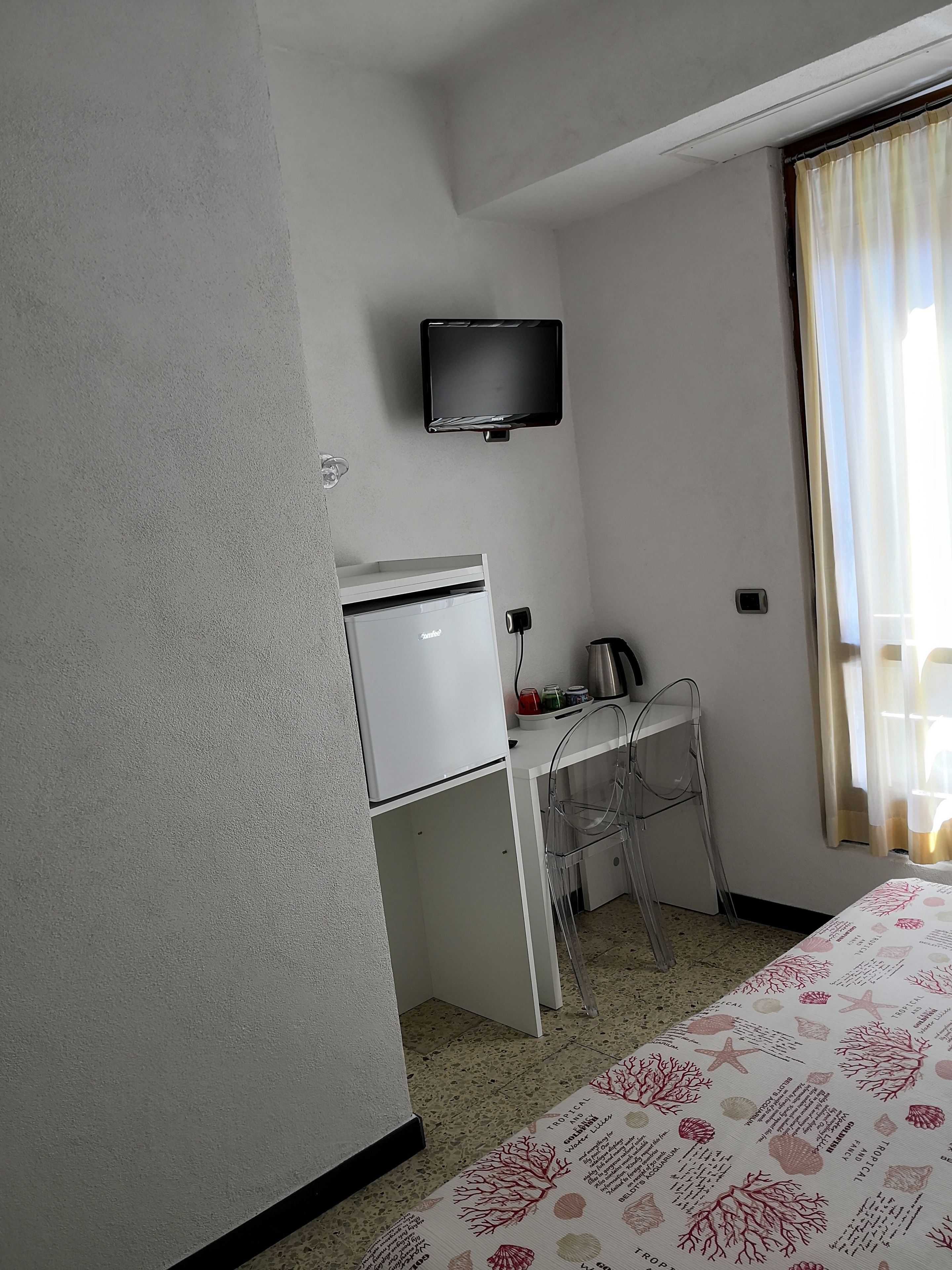 Quarto de hotel compacto com uma cama, escrivaninha com computador, televisão na parede e janela com cortinas.