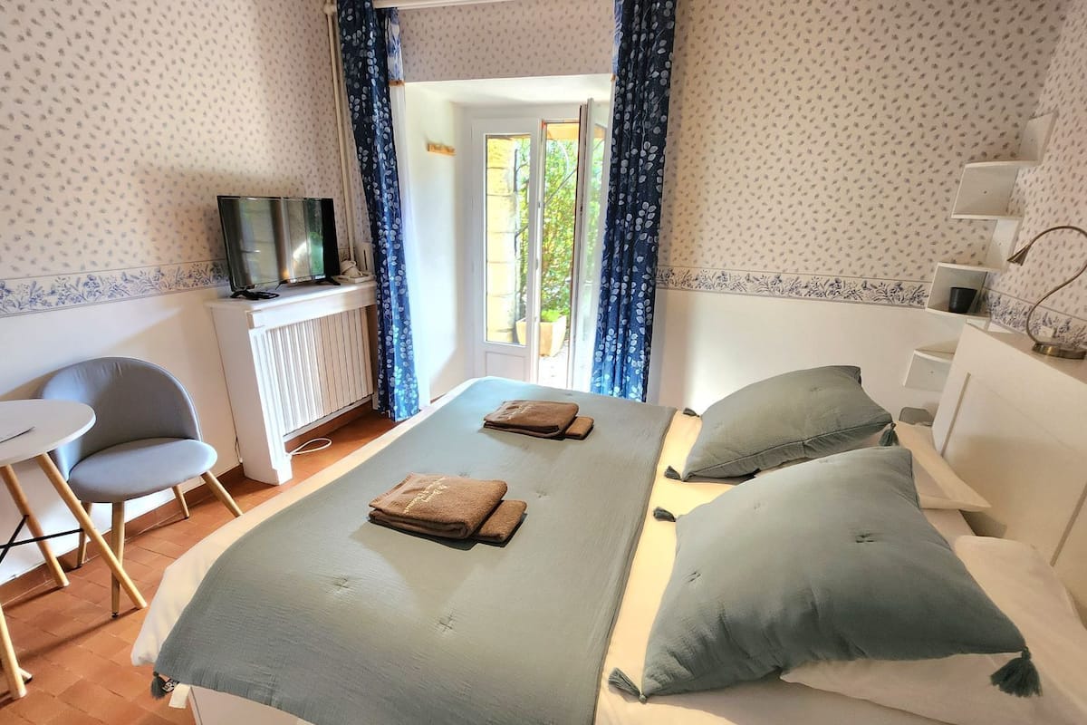 Quarto de hotel com uma cama, dois travesseiros, uma cadeira, uma mesa pequena, televisão e uma janela com cortinas.