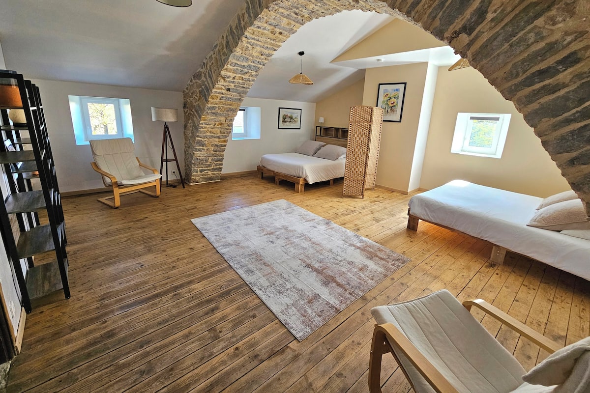 Um quarto de loft aconchegante com um arco de pedra, piso de madeira, uma cama, um sofá, uma poltrona e uma pequena janela.