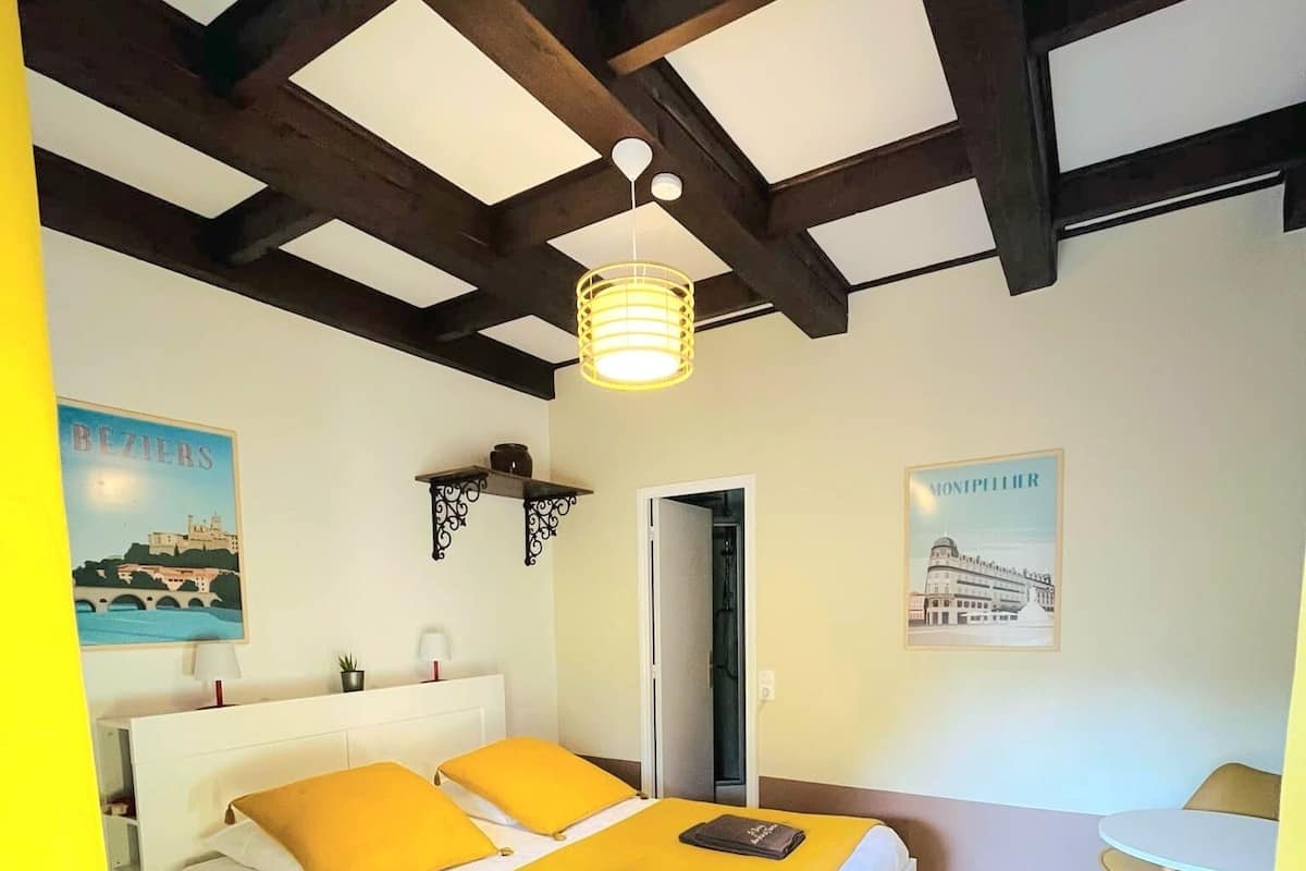 Quarto com cama, mesa de cabeceira, cadeira, luminária pendente e duas imagens penduradas na parede.