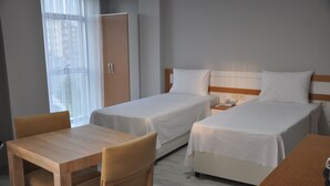Quarto de hotel com duas camas de solteiro, uma escrivaninha de madeira, uma cadeira, um guarda-roupa e uma janela com cortinas leves.