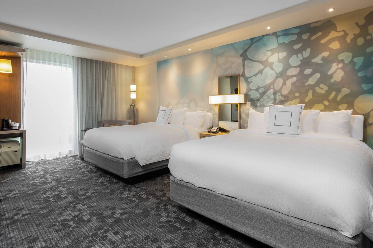 Quarto de hotel com duas camas, uma escrivaninha com luminária, uma cadeira e um grande mural abstrato na parede.