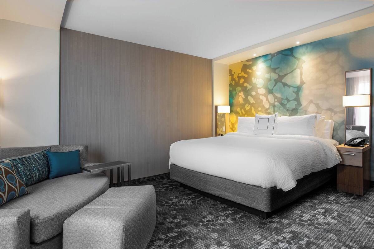 Quarto de hotel moderno com uma cama grande, um sofá, uma mesinha e uma parede com um design abstrato colorido.