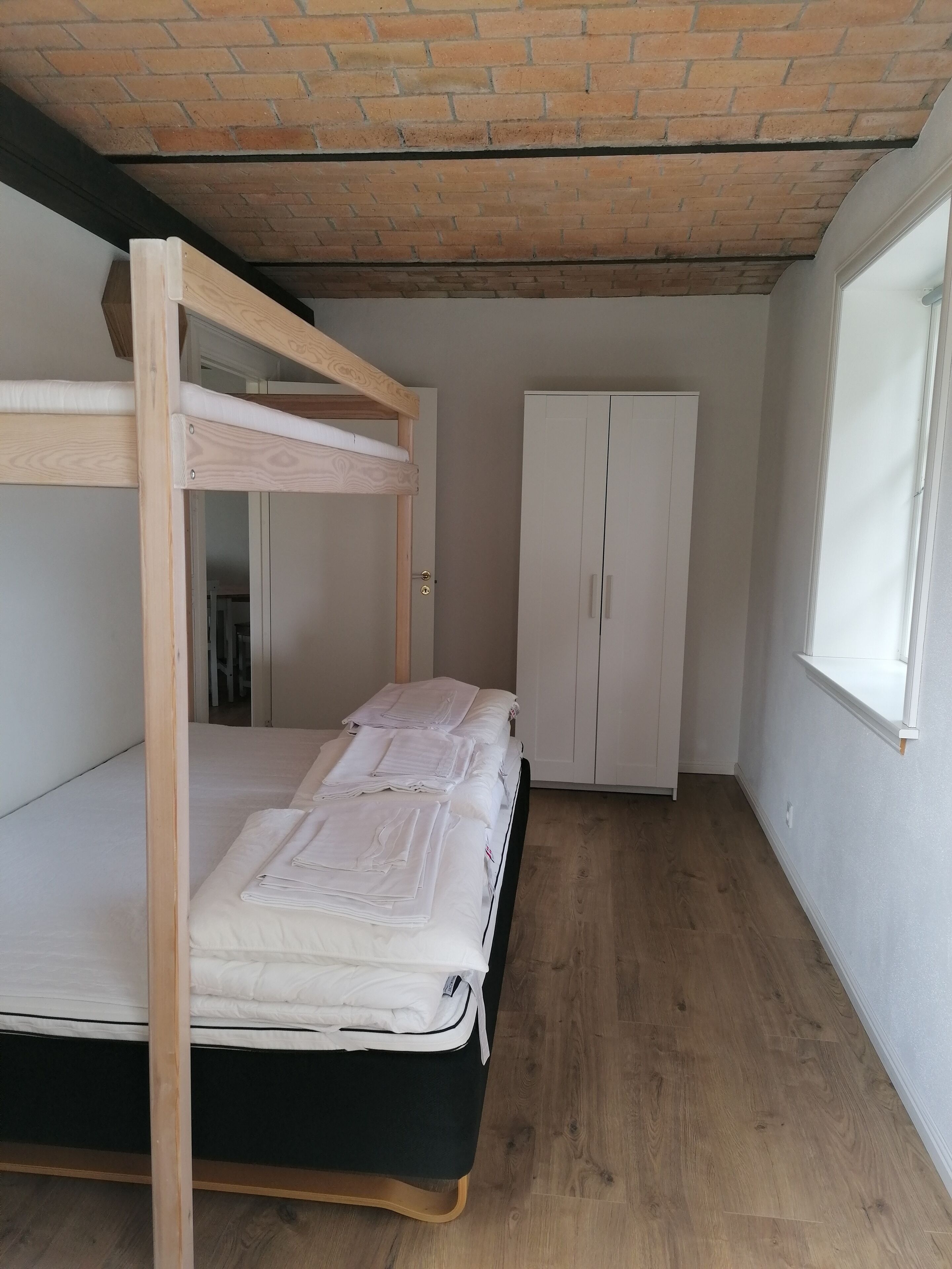 Uma cama beliche com roupa de cama branca, um guarda-roupa branco e uma janela com luz natural.