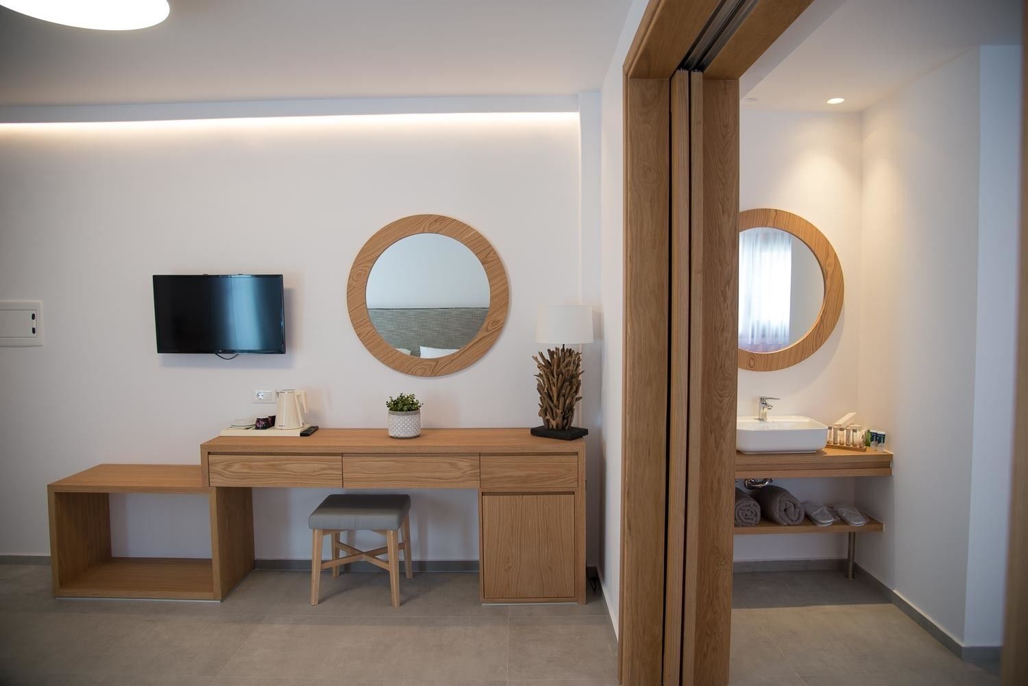 Um quarto moderno com escrivaninha de madeira, televisão, duas espelhos redondos, um banco e mesa de cabeceira com abajur e planta.
