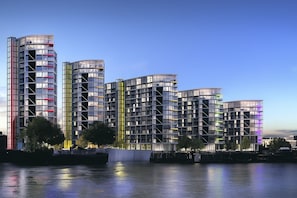 Um edifício moderno alto com várias varandas e fachadas de vidro refletivas, localizado à beira de um corpo d'água.