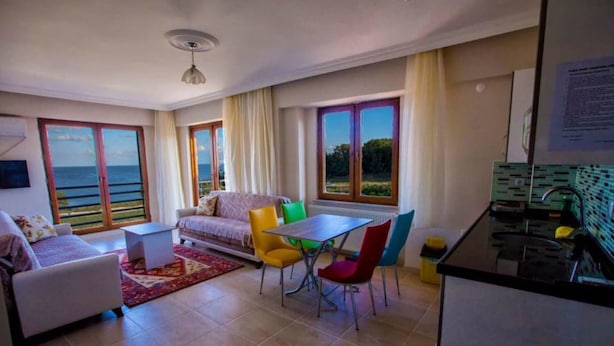 Uma sala de estar moderna com um sofá, uma mesa de jantar com cadeiras coloridas e grandes janelas com vista para o mar.
