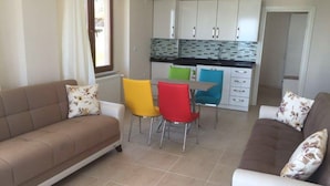 Uma sala de estar com sofá e poltronas, uma mesa de jantar e uma cozinha com armários e painel de azulejos.