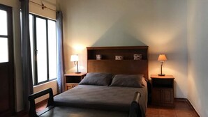 Quarto com cama, duas mesas de cabeceira com luminárias, cabeceira de madeira e um banco em frente à cama.