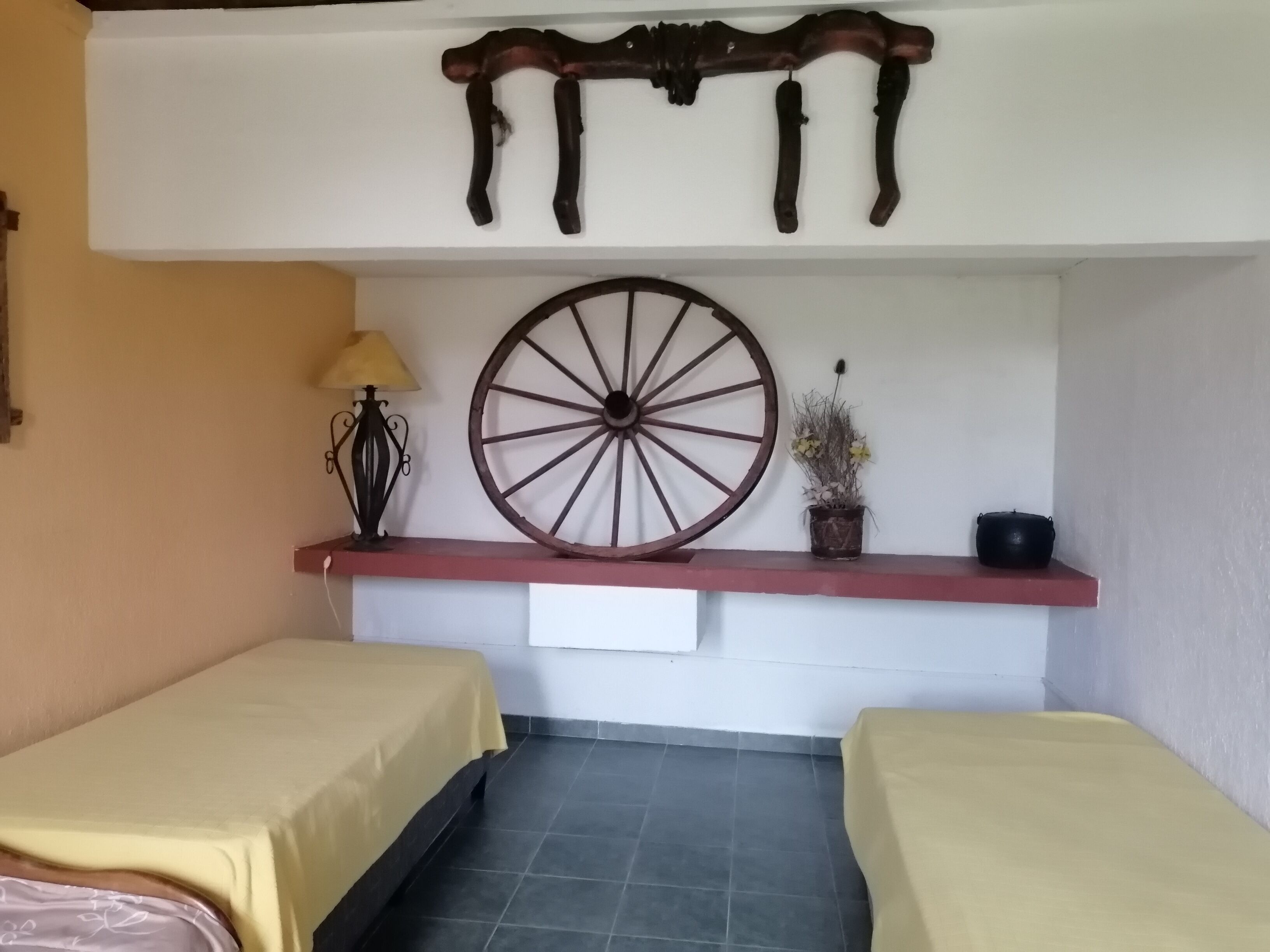 Quarto com duas camas, prateleira de madeira fixada na parede com uma luminária, uma grande roda de madeira e um vaso com planta.