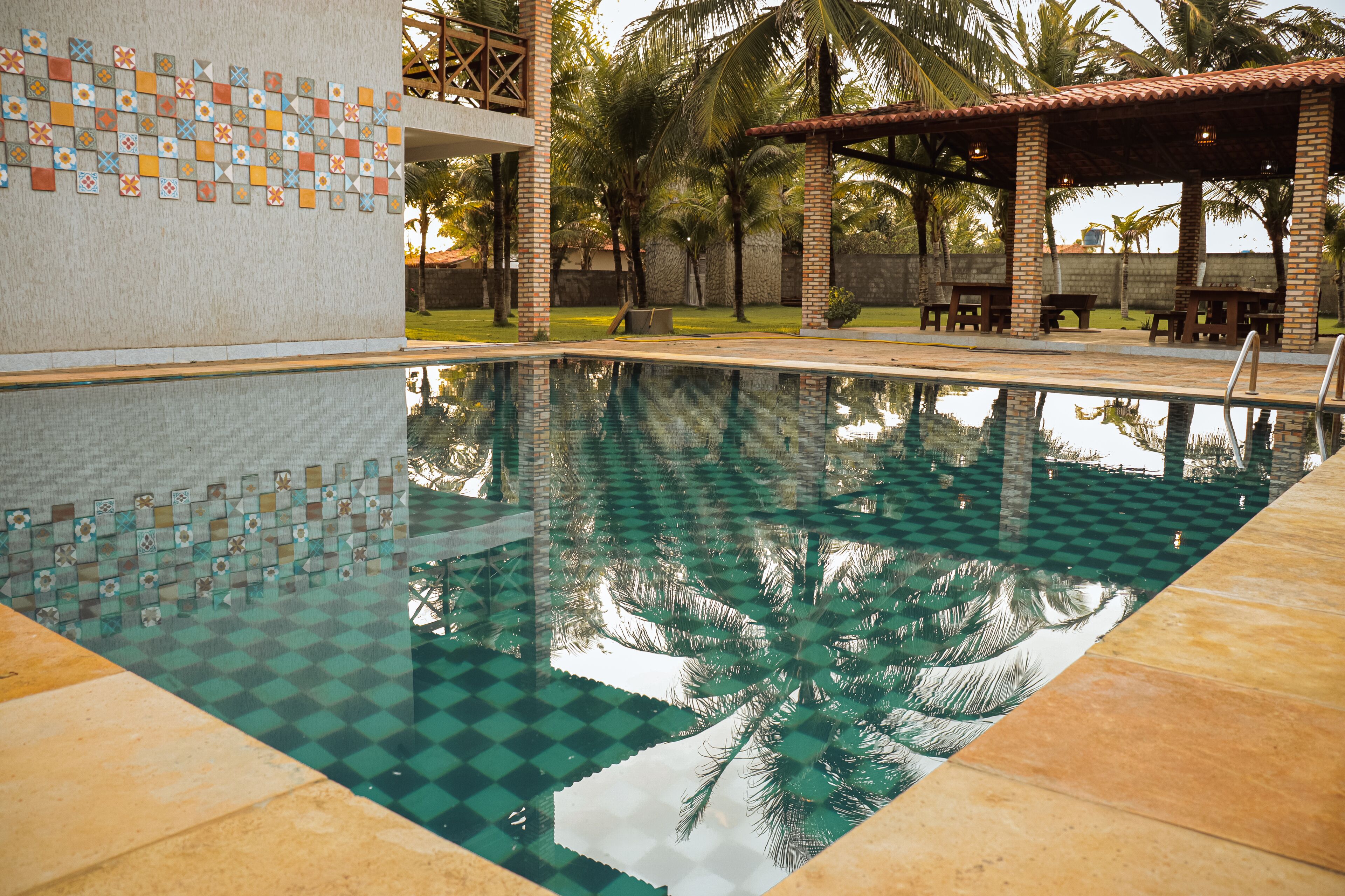 Uma piscina com padrão de azulejos, cercada por palmeiras e uma área coberta com assentos.
