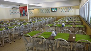 Uma cafeteria com fileiras de mesas e cadeiras, uma televisão na parede e obras de arte nas paredes.
