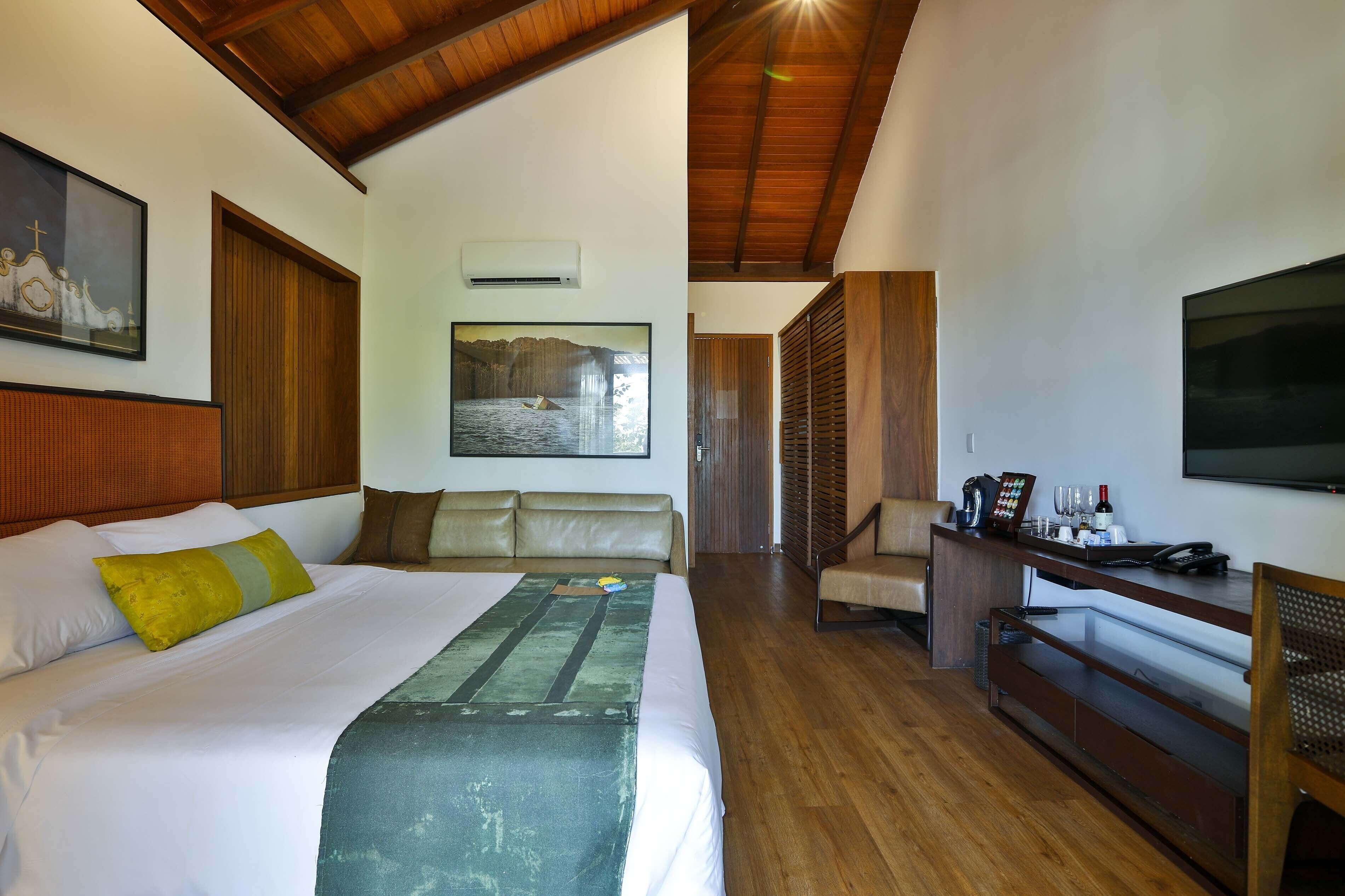 Quarto de hotel com uma cama grande, uma mesa de trabalho com telefone e óculos, e uma televisão na parede.