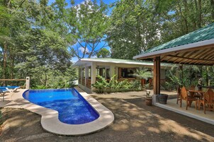 Uma piscina com borda curva, rodeada por vegetação exuberante e uma área de estar coberta ao ar livre com mobiliário de madeira.