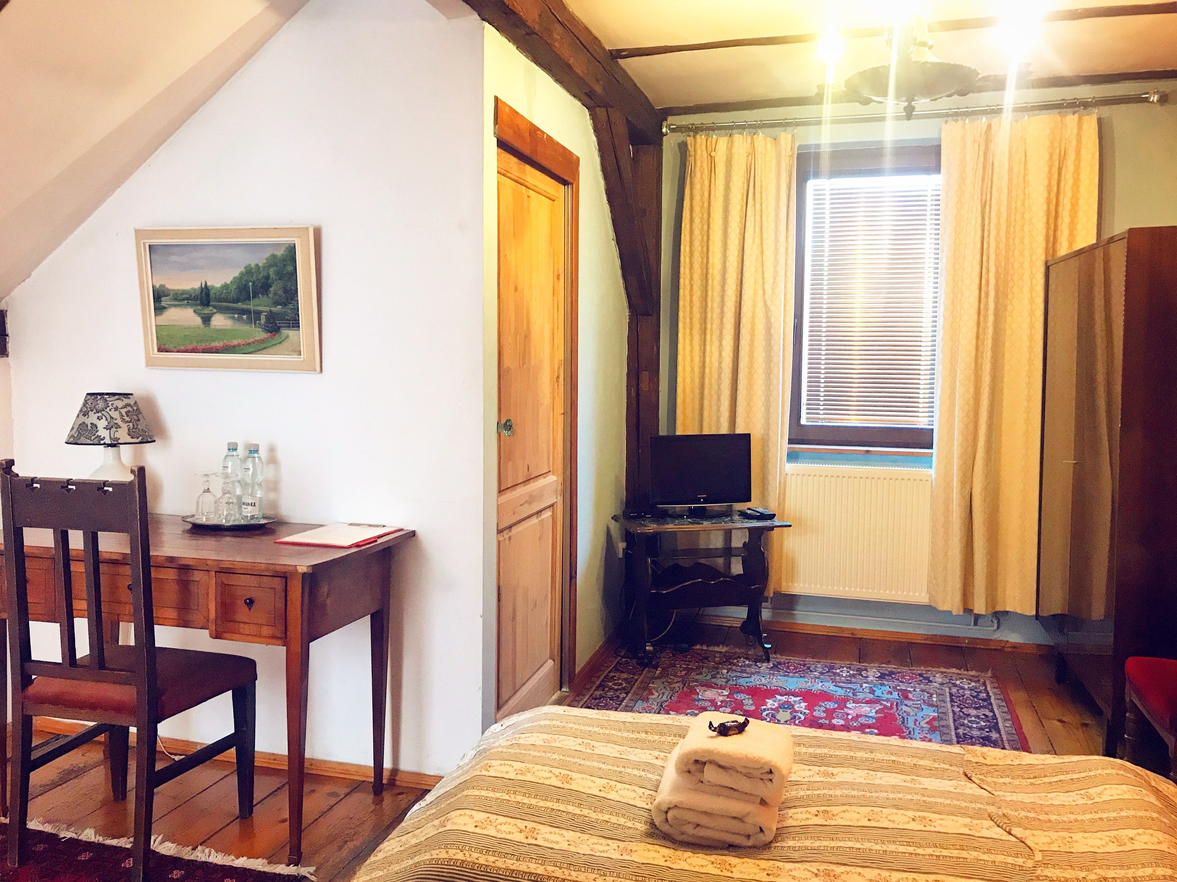 Quarto com cama, escrivaninha, cadeira, televisão e janela com cortinas.