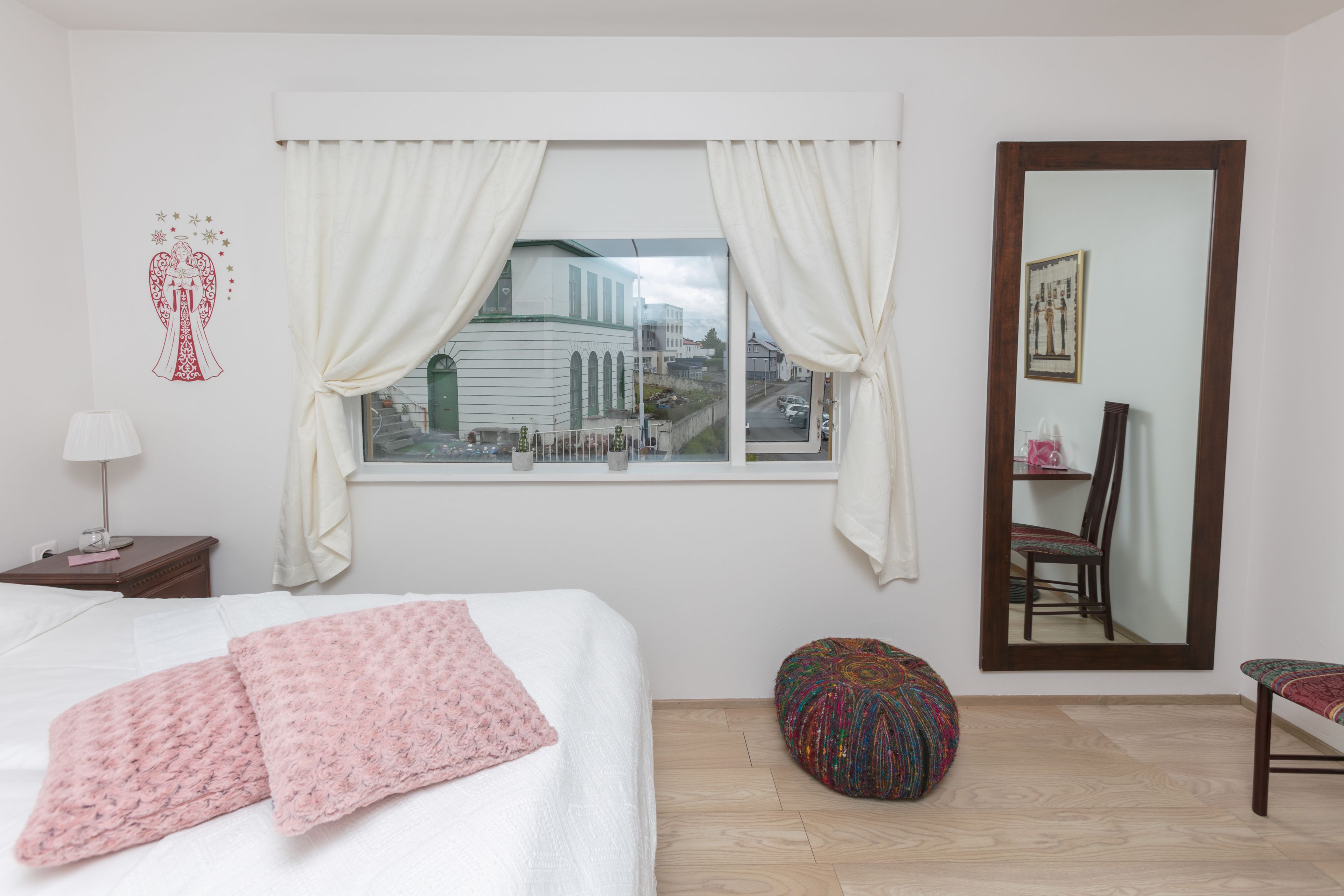 Quarto com cama, janela com cortinas, espelho, cadeira e uma pequena mesa com abajur.