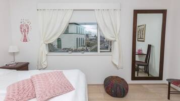 Quarto com cama, janela com cortinas, espelho, cadeira e uma pequena mesa com abajur.