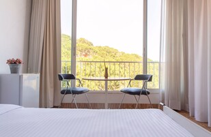 Quarto com cama, duas cadeiras, uma mesinha e janela com vista para área verde.