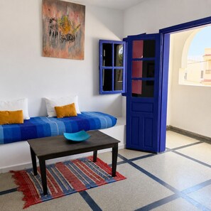 Quarto com sofá azul, mesa pequena, tapete colorido, porta azul e um quadro na parede.