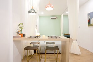 Uma sala de jantar com uma mesa posta para quatro pessoas, duas cadeiras, uma planta em vaso e um quadro na parede.