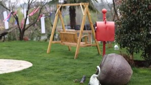 Um jardim com balanço de madeira, caixa de correio vermelha e uma grande pedra decorativa.