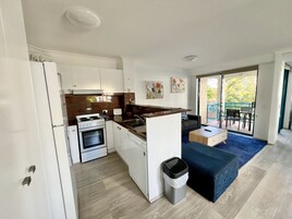 Uma cozinha compacta com fogão, forno e pia, uma área de jantar com mesa e cadeiras, e uma varanda com vista para o verde.