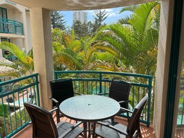 Uma varanda com mesa redonda e quatro cadeiras, cercada por folhagem exuberante e vista para edifícios altos ao fundo.