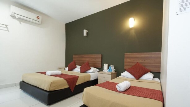 Quarto de hotel com duas camas, cada uma com cabeceira de madeira, roupa de cama bege e almofadas decorativas vermelhas. Há uma mesa de cabeceira com abajur e artigos de higiene, e um aparelho de ar condicionado na parede.