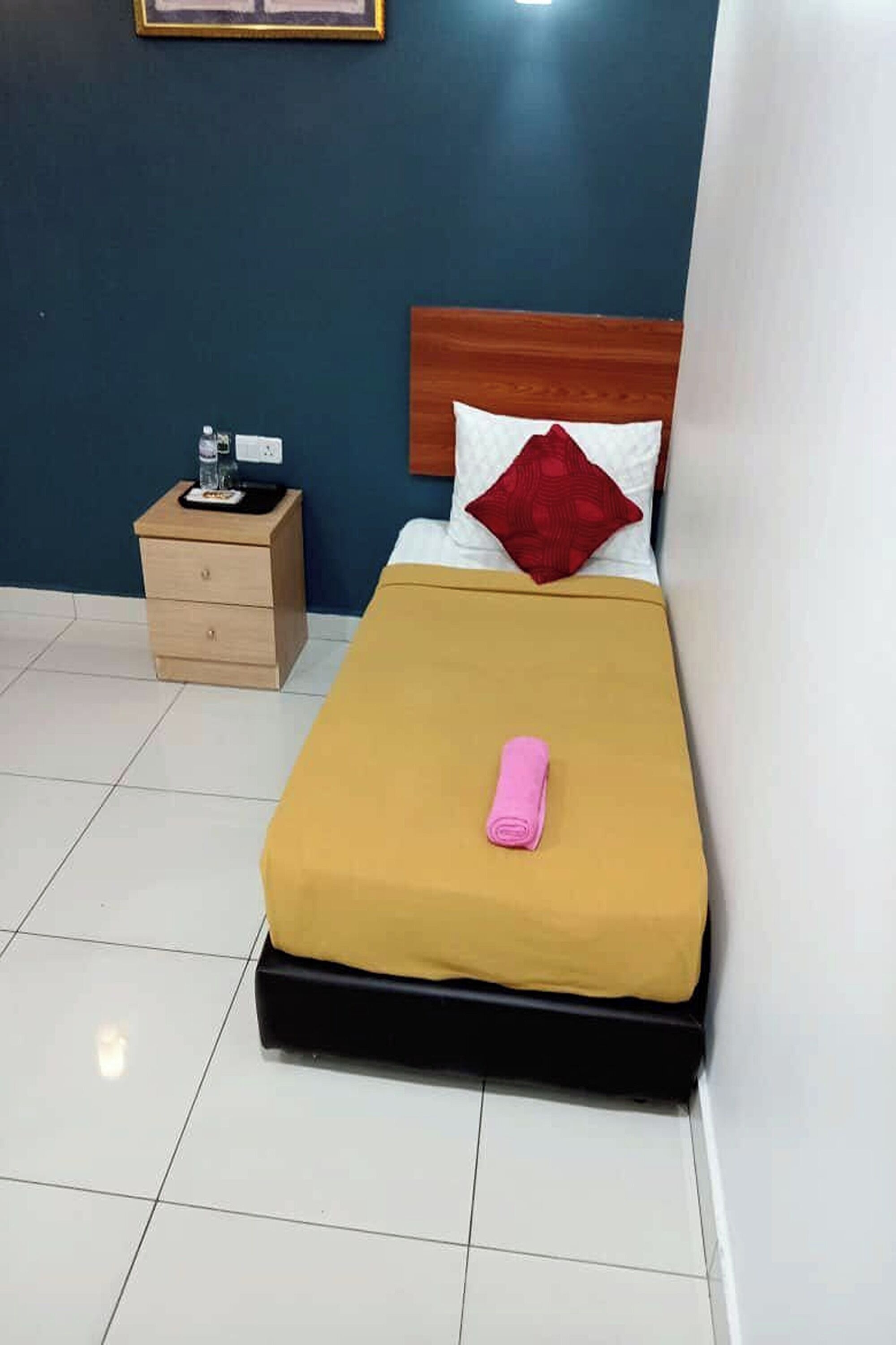 Uma cama bem arrumada com colcha amarela e toalha rosa, uma mesa de cabeceira de madeira com garrafa e copos, e um travesseiro vermelho na cama.