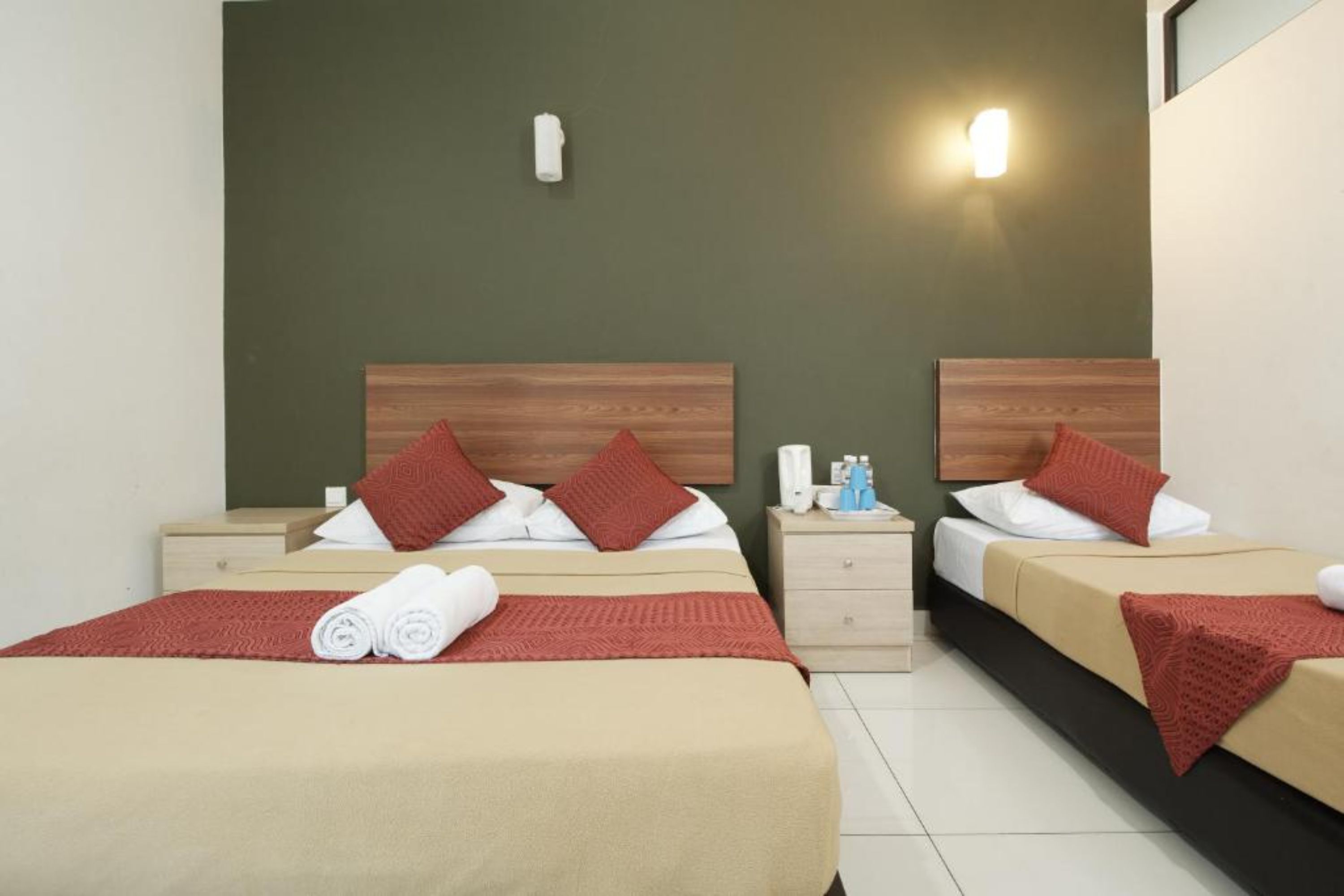 Quarto de hotel com duas camas, cada uma com cabeceira de madeira, almofadas vermelhas e brancas e um cobreleiro bege. Há duas mesas de cabeceira com luminárias e uma pequena mesa com produtos de higiene pessoal.