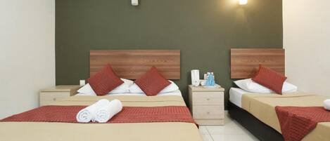 Quarto de hotel com duas camas, cada uma com cabeceira de madeira, almofadas vermelhas e brancas e um cobreleiro bege. Há duas mesas de cabeceira com luminárias e uma pequena mesa com produtos de higiene pessoal.