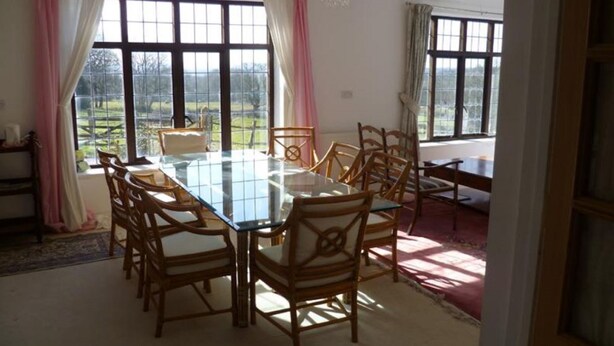 Uma sala de jantar com mesa de vidro e cadeiras de madeira, janelas amplas com cortinas e vista para um jardim.