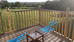 Um deck de madeira com mesa de vidro e duas cadeiras azuis, com vista para uma paisagem verde.