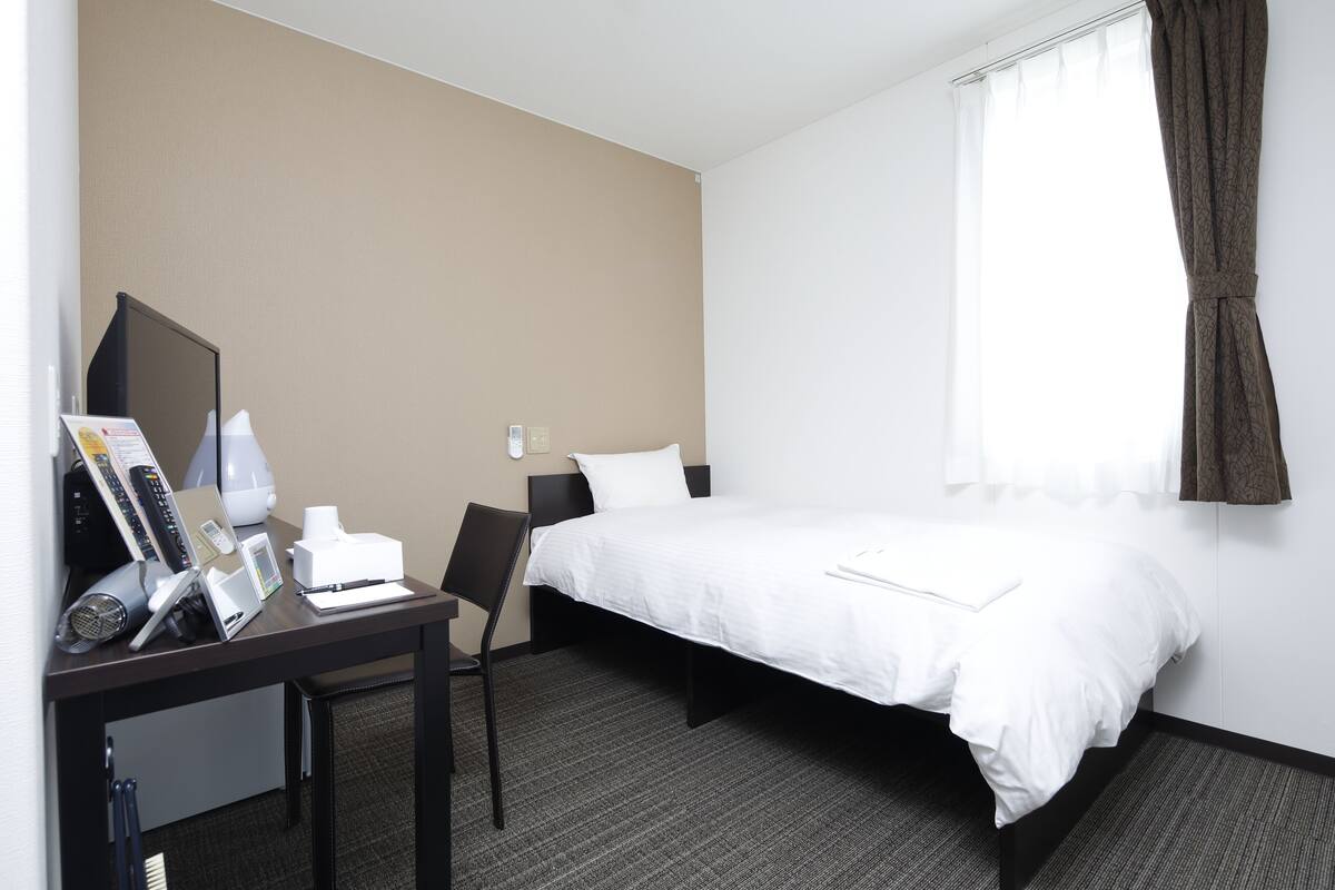 Quarto de hotel com cama, escrivaninha com cadeira, telefone e janela com cortinas.