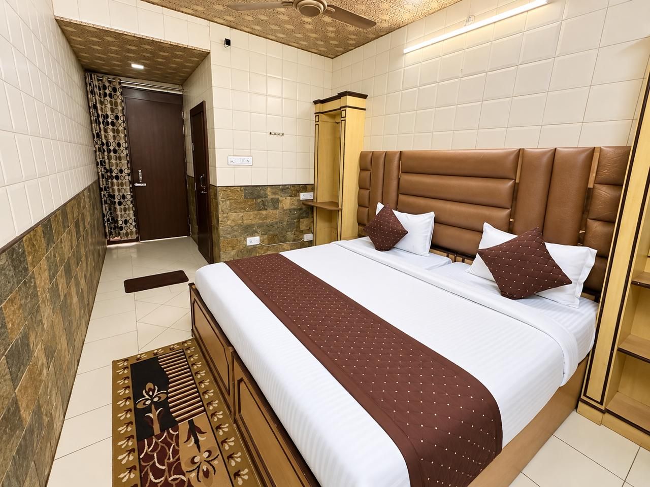 Quarto de hotel com cama grande, cabeceira de madeira, tapete estampado e parede decorativa em pedra.
