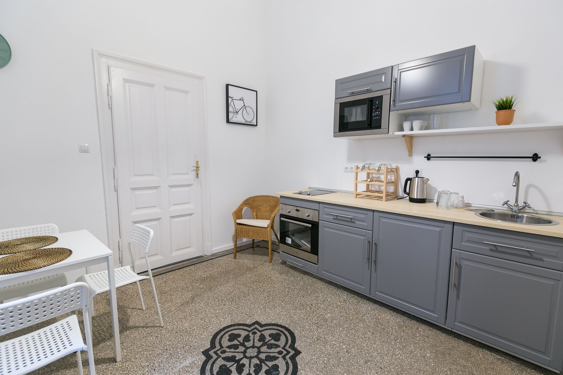 Uma cozinha compacta com armários cinza, micro-ondas e pia. Há uma cadeira de vime e uma mesa branca com jogos americanos trançados.