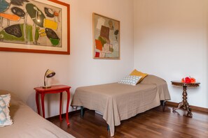 Quarto com duas camas de solteiro, uma pequena mesa vermelha com abajur, piso de madeira e duas pinturas nas paredes.