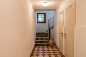 Um corredor estreito com chão quadriculado, uma janela no topo de uma escada e uma porta à direita.