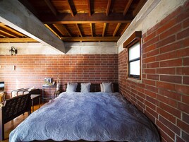 Quarto com parede de tijolos, vigas de madeira no teto, cama com colcha estampada, mesa pequena com abajur e uma janela.