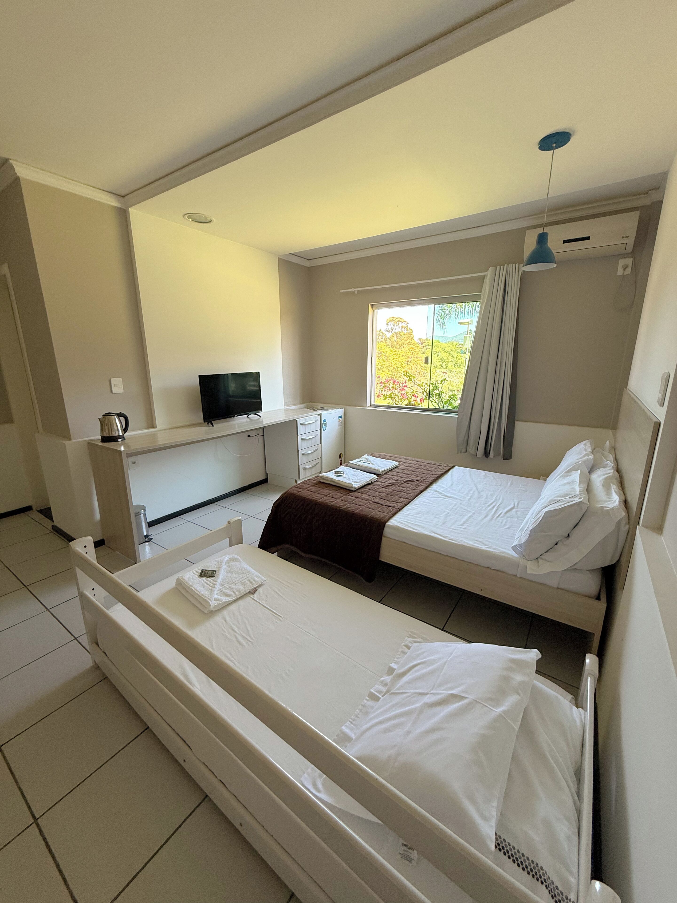 Quarto de hotel com cama, escrivaninha, televisão e janela com cortinas.