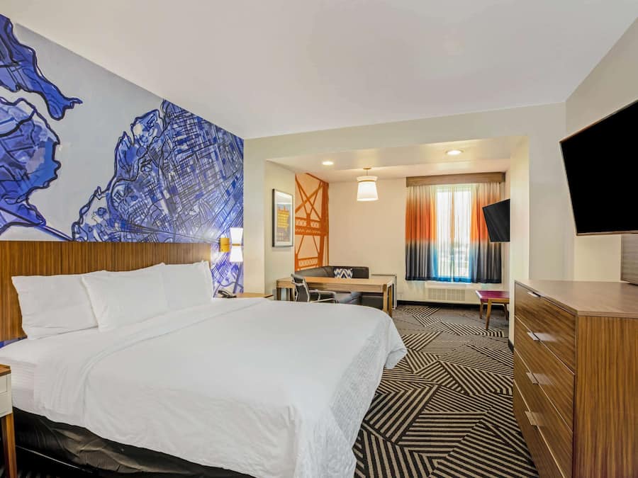 Quarto de hotel com uma cama grande, uma escrivaninha, uma cadeira, uma televisão e um tapete estampado.
