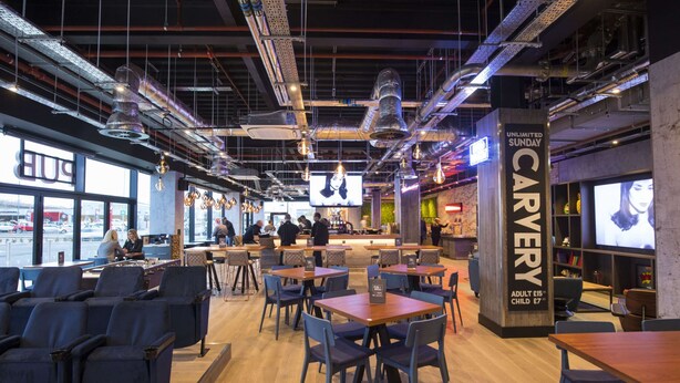 Interior de restaurante moderno com diversas áreas de estar, um bar e grandes telas exibindo conteúdo.